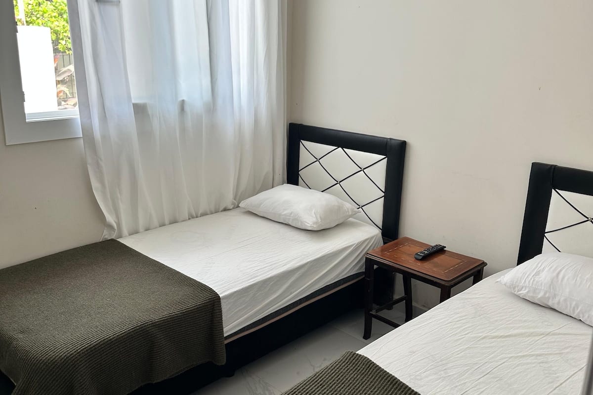 Quarto de hotel com duas camas de solteiro, uma pequena mesa de cabeceira de madeira, um aparelho de ar condicionado e uma janela com cortinas brancas.