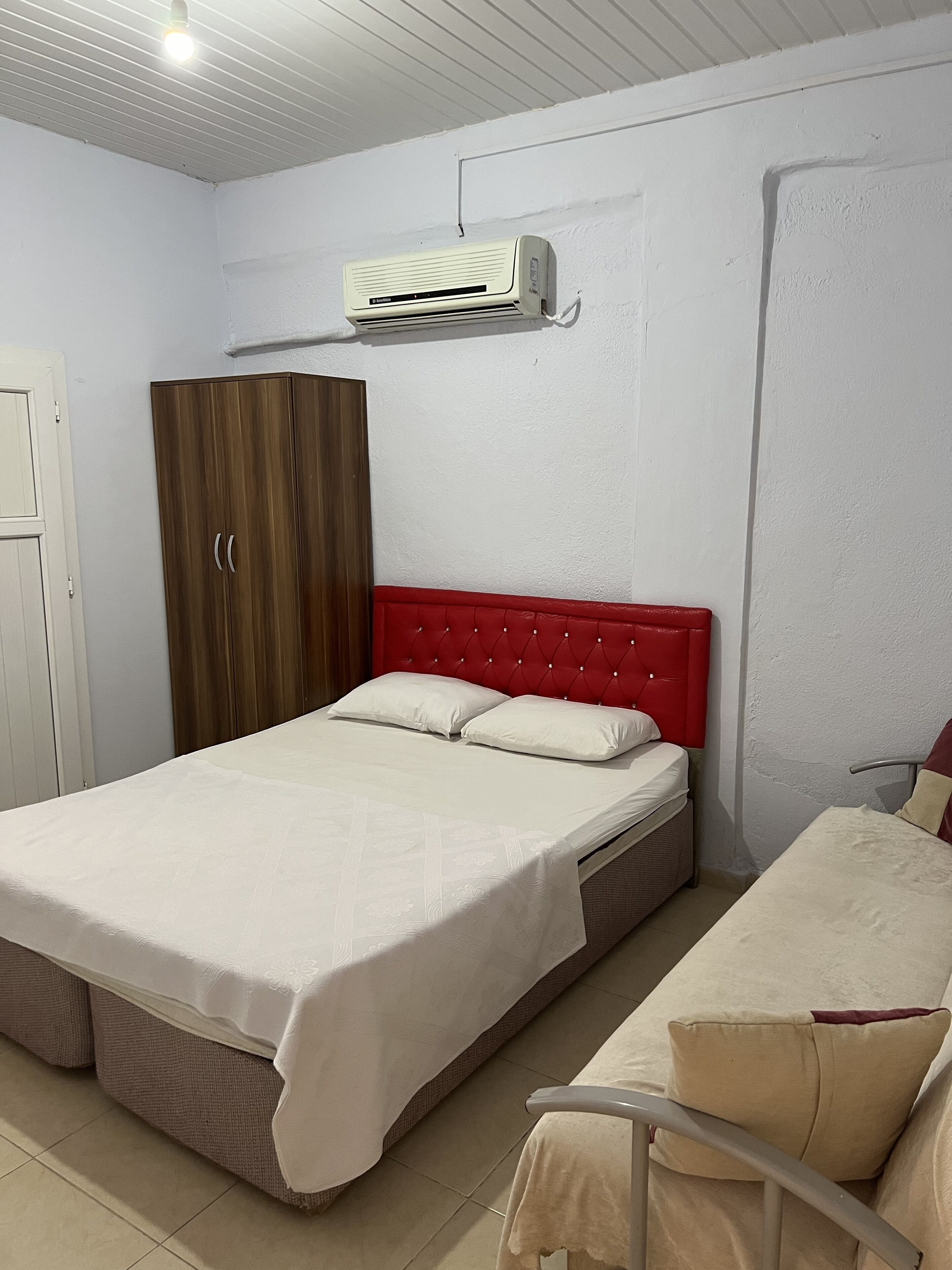 Quarto com cama, guarda-roupa, cabeceira estofada vermelha e ar condicionado na parede.