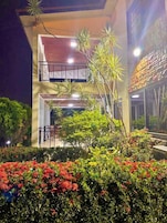 Um edifício de dois andares com varanda, cercado por vegetação exuberante e flores vibrantes.