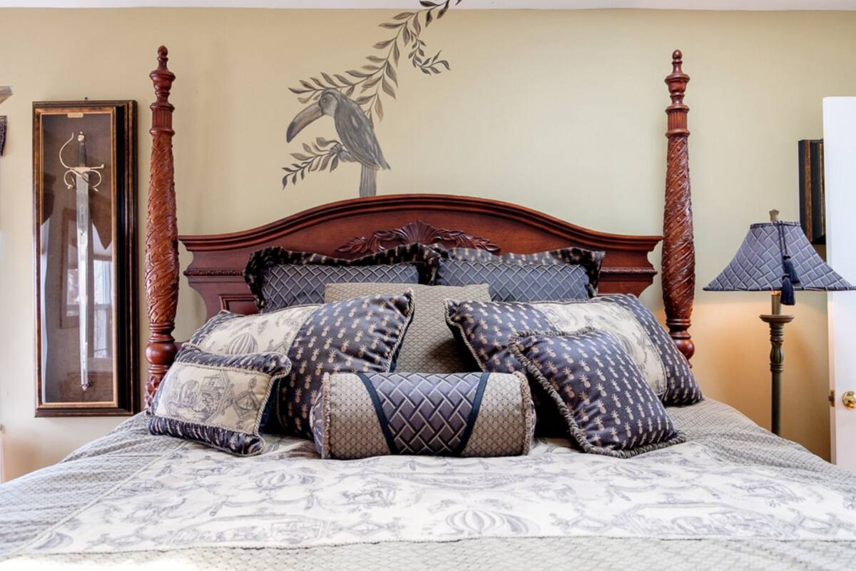 Uma cama com colcha estampada e várias almofadas, uma obra de arte na parede com um pássaro e um abajur na lateral.