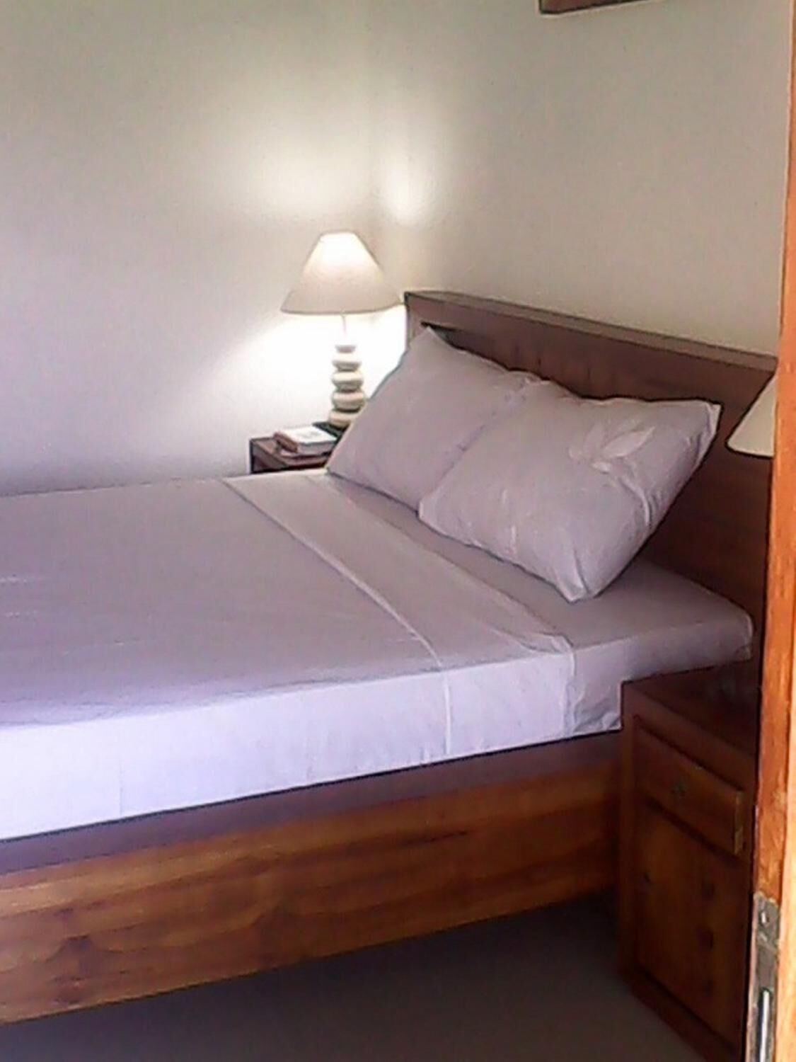 Uma cama de casal com cabeceira de madeira, uma mesa de cabeceira com abajur e um criado-mudo com livros.