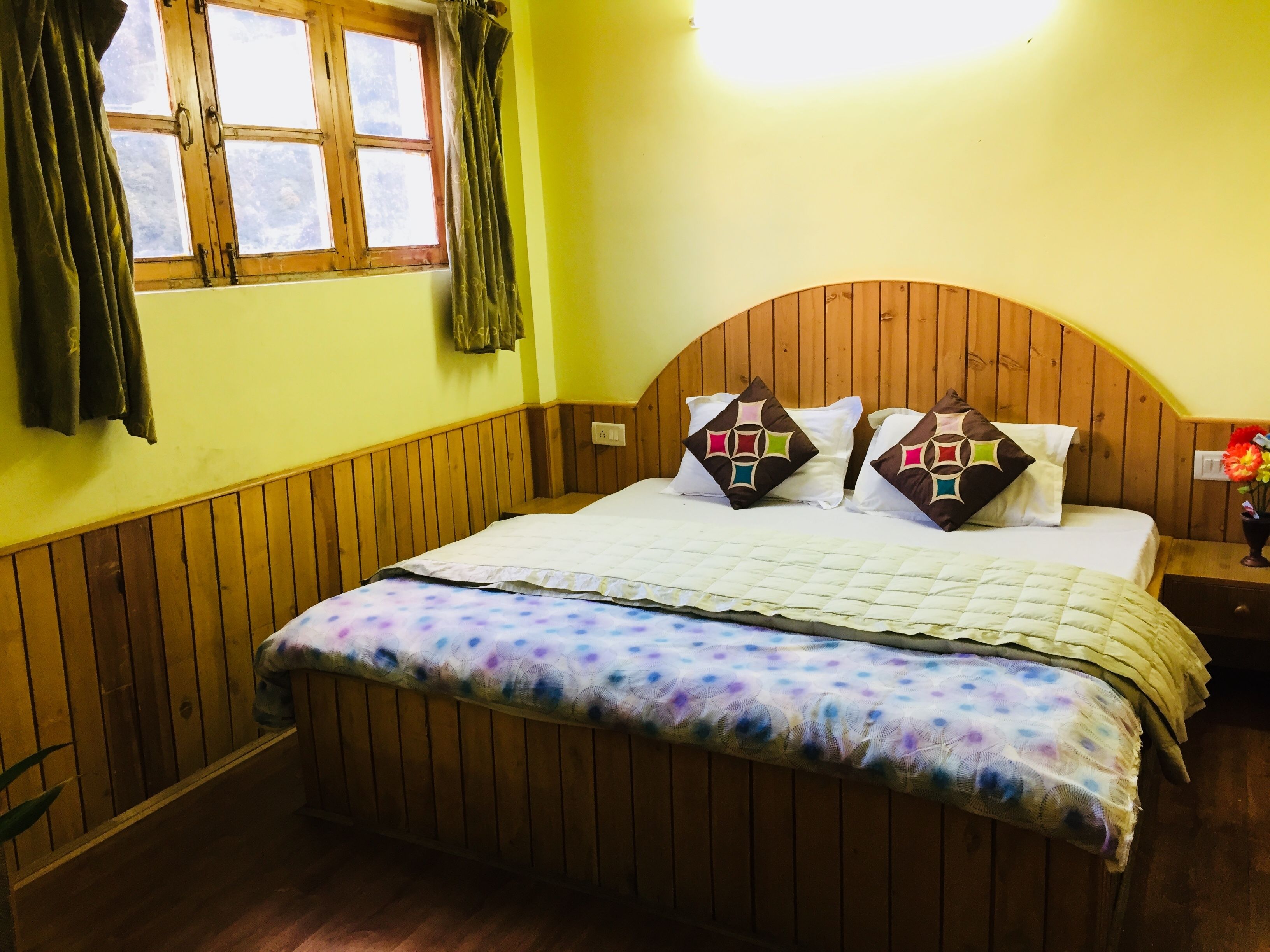 Uma cama com colcha, duas almofadas, uma mesa de cabeceira, uma janela com cortinas e painéis de madeira nas paredes.