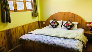 Uma cama com colcha, duas almofadas, uma mesa de cabeceira, uma janela com cortinas e painéis de madeira nas paredes.