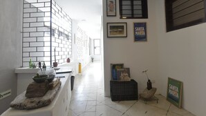 Um corredor com piso de azulejos, parede branca com padrão de tijolos e um balcão com uma tigela e uma garrafa. Há quadros emoldurados e uma placa na parede.