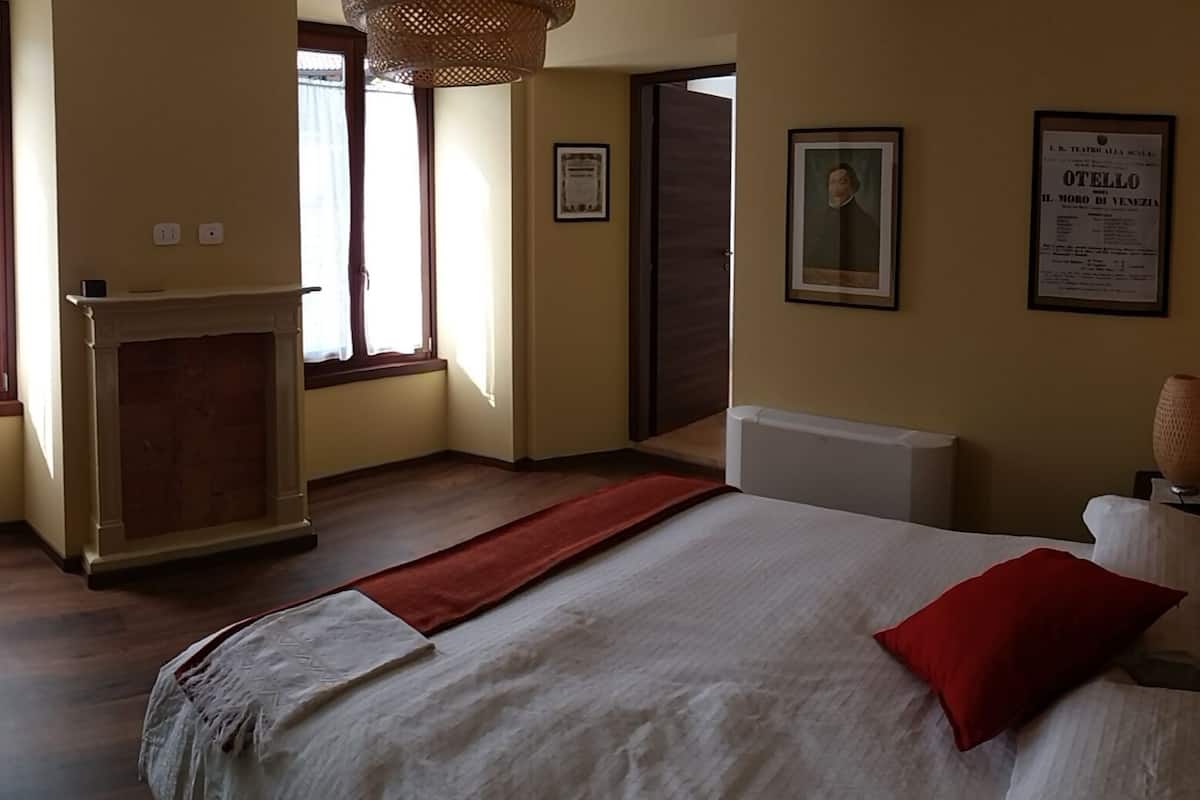 Quarto de hotel com cama, lareira, janela e parede com um quadro emoldurado e um cartaz.