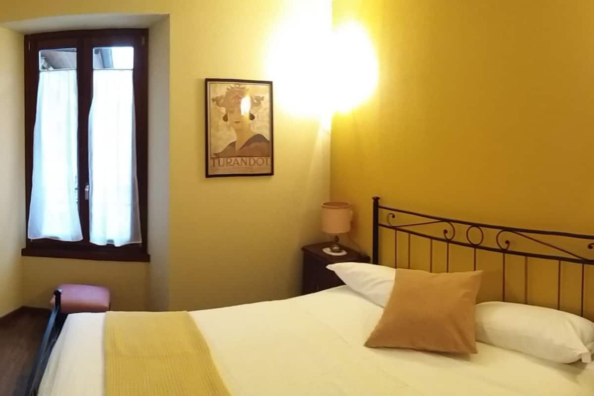 Quarto com cama, duas luminárias de cabeceira, janela com cortinas e um pôster na parede.