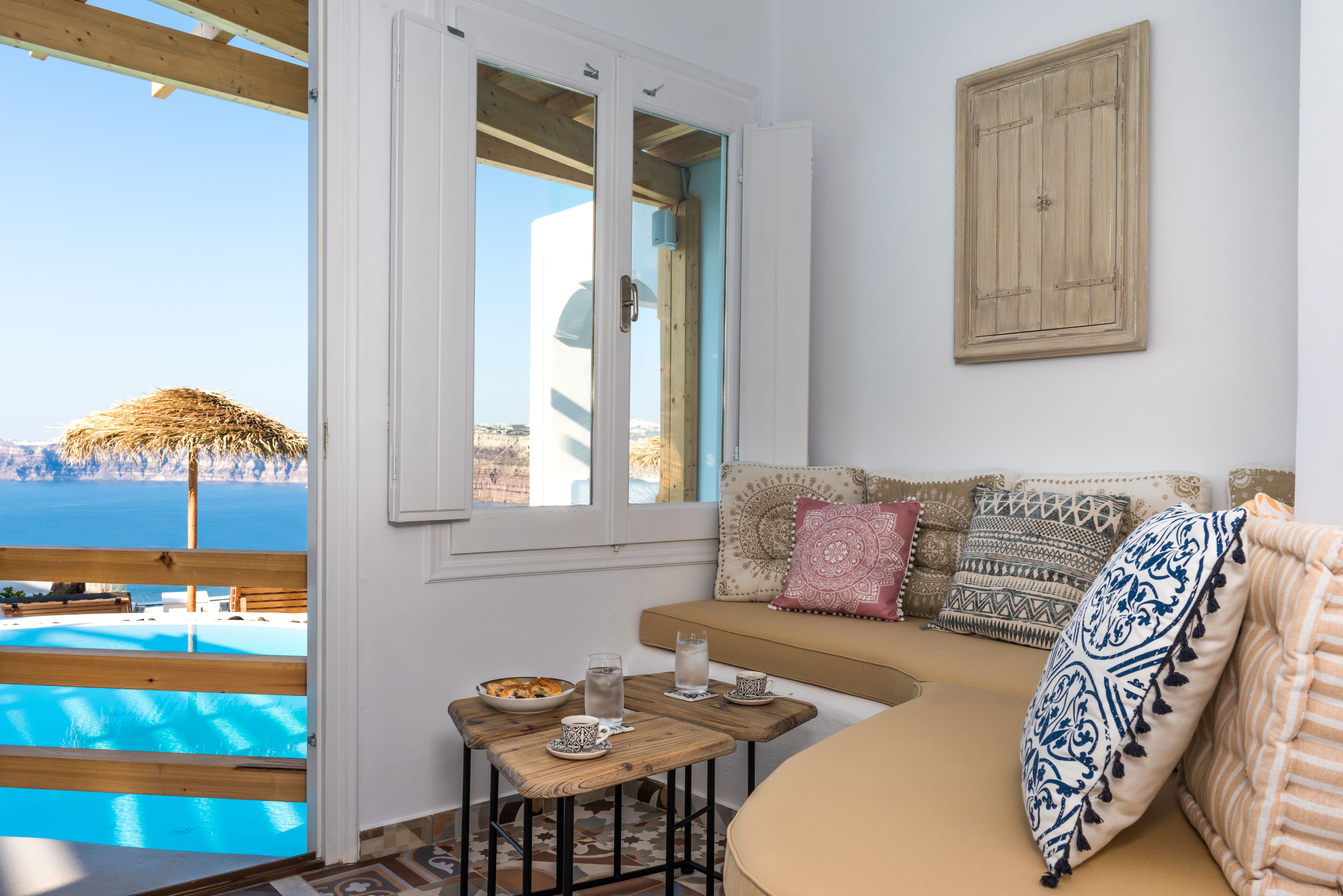 Quarto com vista para uma piscina e o mar, uma mesa de madeira com uma tigela e copos, e um sofá com almofadas estampadas.