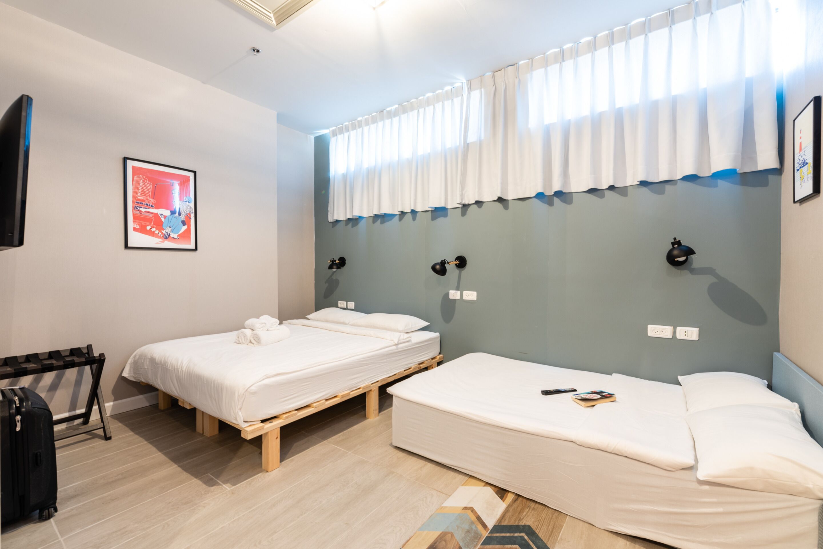 Quarto de hotel com duas camas de solteiro, um banco com uma mala e um quadro na parede.
