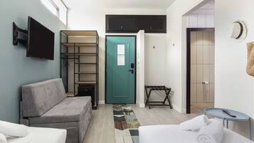 Quarto de hotel compacto com sofá, cama, televisão na parede, porta verde-água e uma pequena mesa com controle remoto.