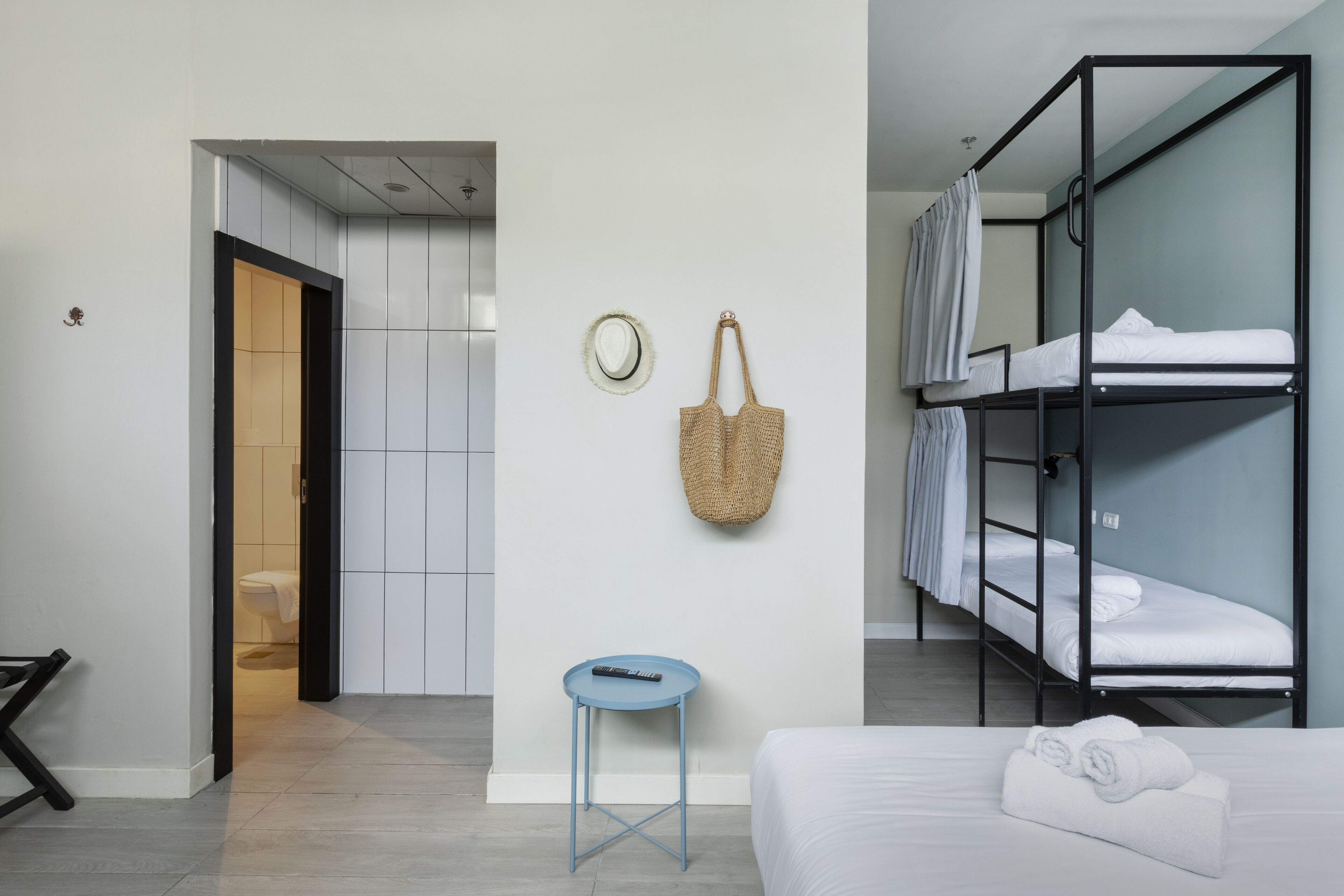 Quarto de hotel com cama beliche, uma pequena mesa redonda, uma bolsa pendurada e uma porta que dá para outro quarto.