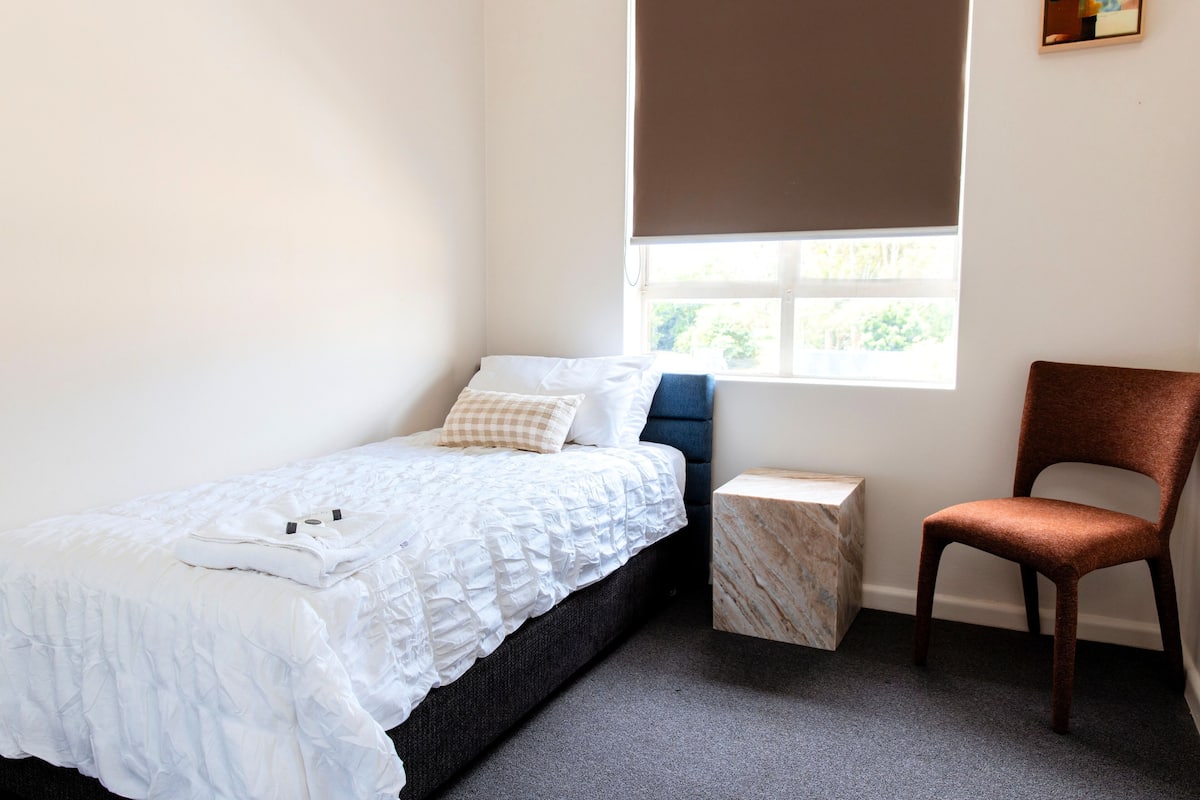Quarto com cama, cadeira, mesa de cabeceira e janela com vista para o verde.