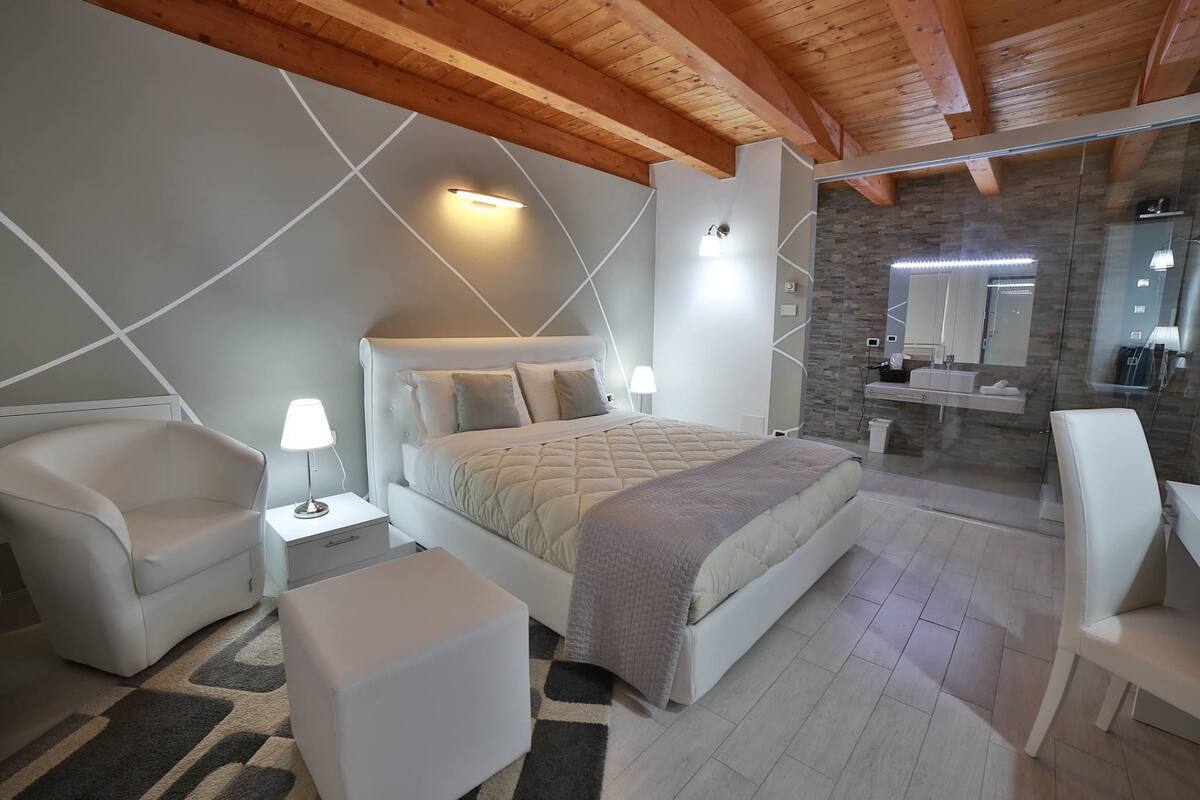 Quarto moderno com uma cama grande, um espaço de estar com uma poltrona e uma mesinha, e um banheiro com paredes de vidro, espelho e pia.