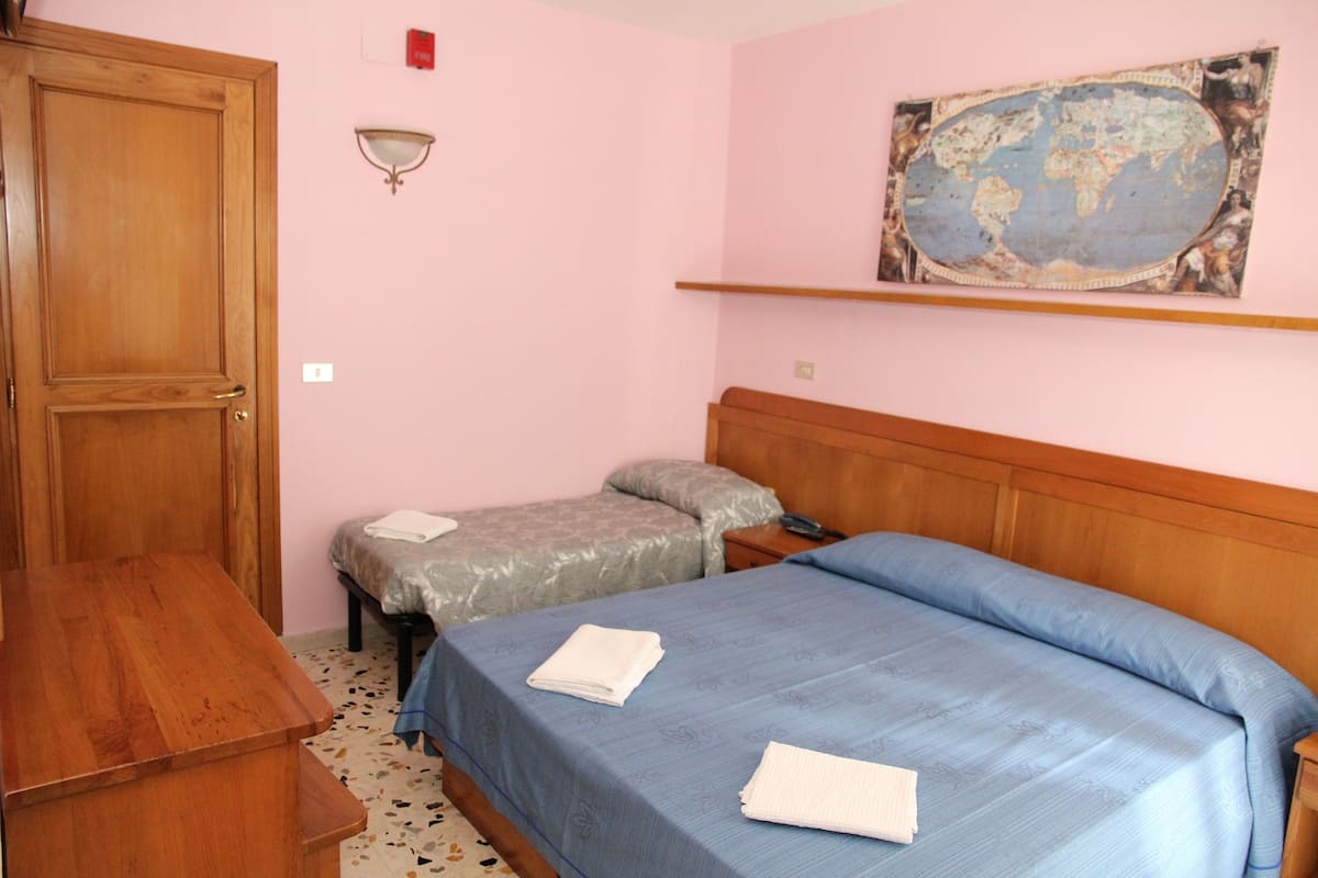 Quarto com duas camas, uma escrivaninha de madeira, guarda-roupa, um mapa-múndi na parede e uma pequena estante.