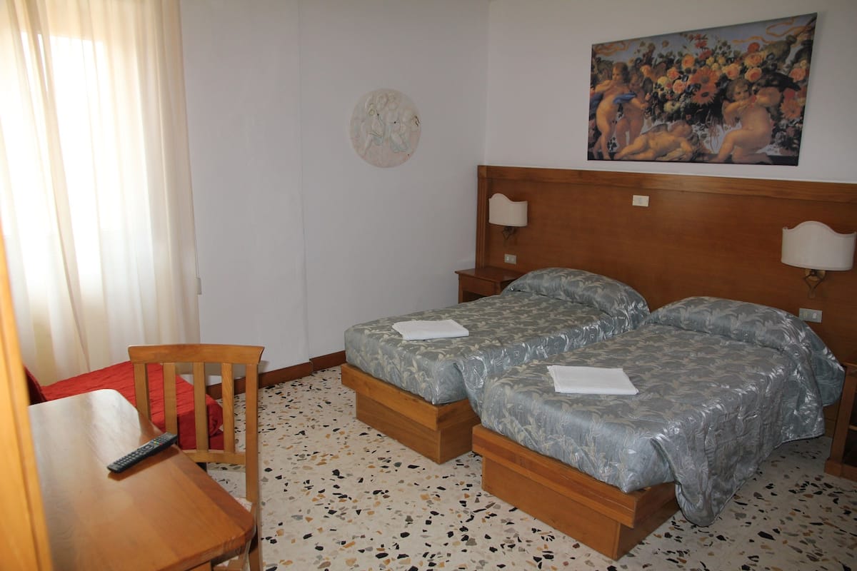 Quarto de hotel com duas camas, cabeceira de madeira, escrivaninha com cadeira, quadro na parede e relógio.