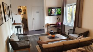 Sala de estar com sofá, mesa de centro e televisão na parede. Cozinha visível ao fundo.