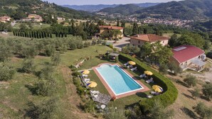 Piscina com espreguiçadeiras e guarda-sóis, rodeada por um gramado bem cuidado e sebes, com vista para uma cidade e montanhas ao fundo.