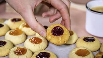 Uma bandeja com biscoitos redondos e geleia no centro, uma mão alcançando um deles, e uma xícara branca ao fundo.
