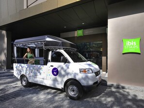 Uma carrinha branca com padrões coloridos de bolinhas estacionada em frente a um edifício com um letreiro verde que diz 'ibis Styles Hotels'.