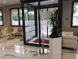 Um hall de hotel com entrada por porta de vidro, cadeiras brancas, uma planta em vaso e tapete vermelho.