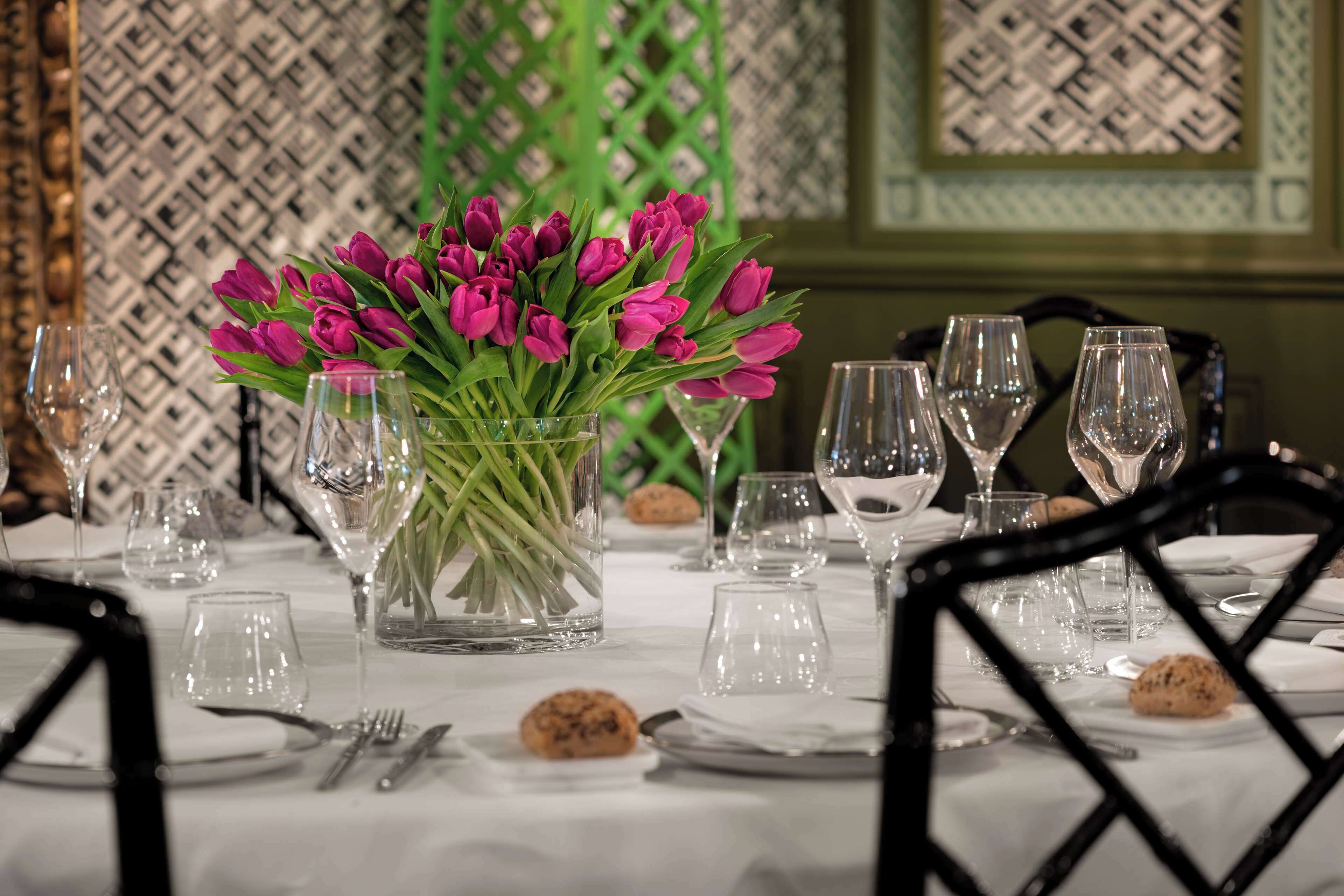 Uma mesa de jantar posta para uma refeição, com um vaso de tulipas cor-de-rosa, taças de vinho e talheres.