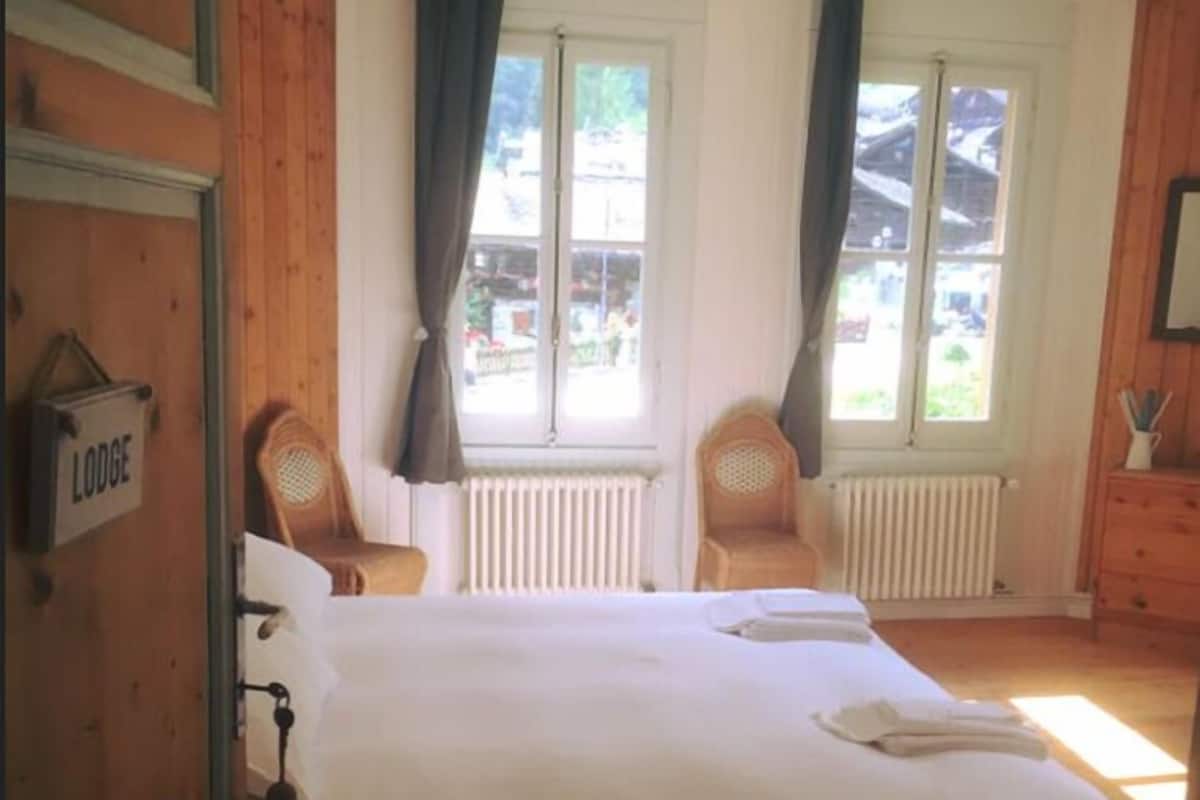 Quarto com uma cama grande, duas cadeiras, uma janela com cortinas e uma placa com a inscrição 'Lodge'.