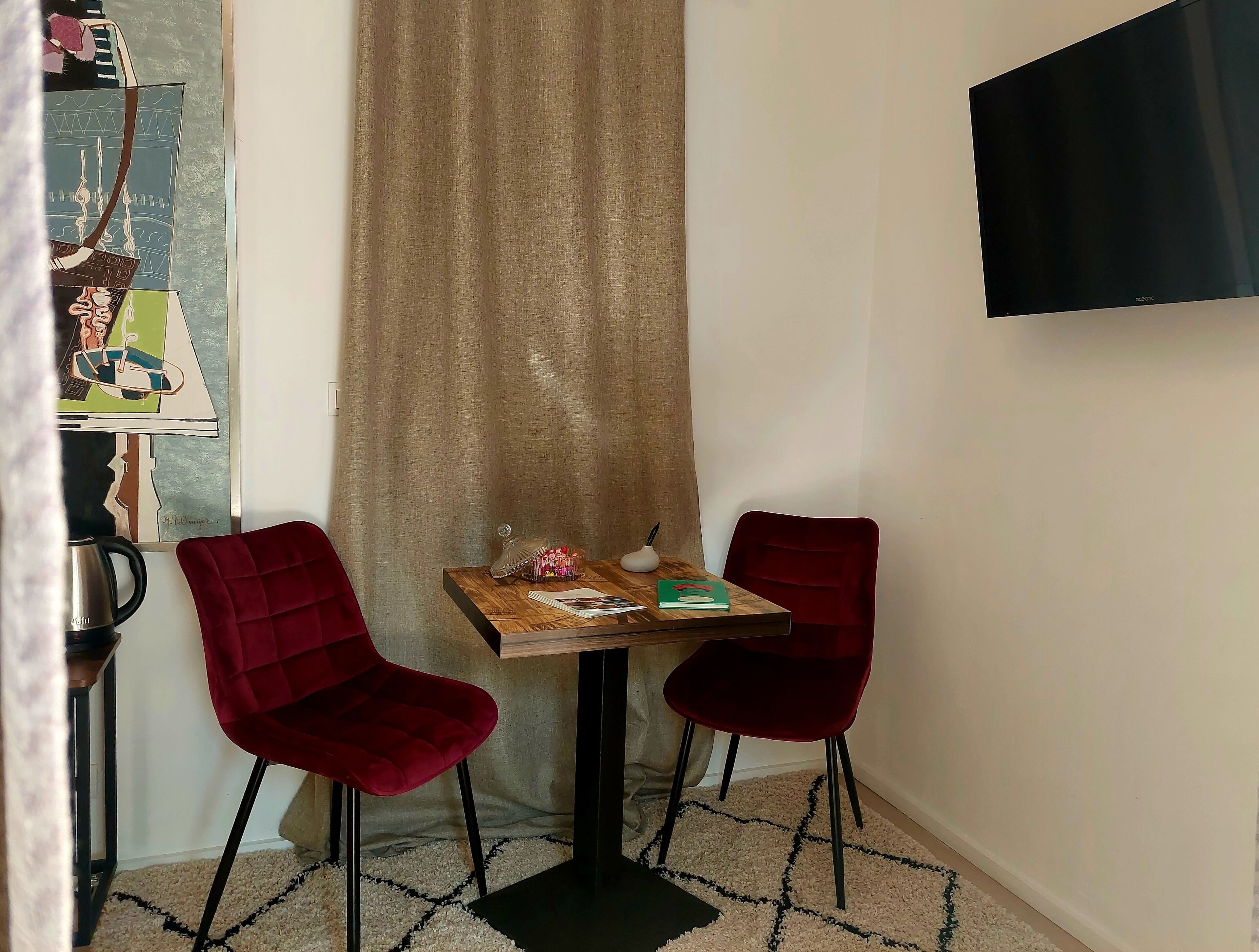 Um quarto pequeno com duas cadeiras vermelhas, uma mesa de madeira, uma televisão e um quadro na parede.