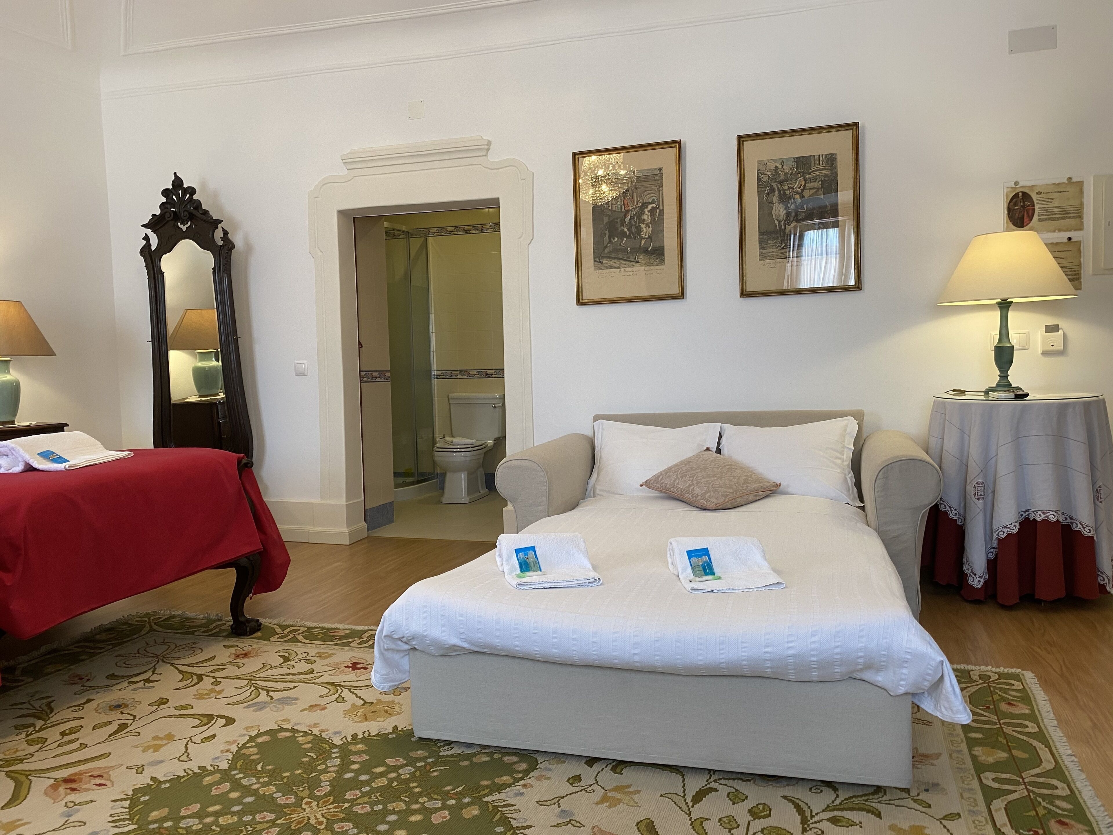 Quarto de hotel com uma cama, uma cadeira vermelha, um banheiro e duas fotos emolduradas na parede.