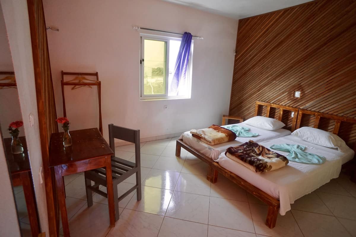 Um quarto com uma cama, cabeceira de madeira, janela com cortina, mesa com um vaso de flores e uma cadeira.