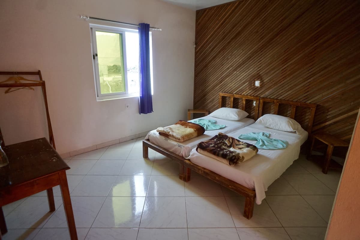 Quarto com duas camas de madeira, uma cadeira de madeira, uma mesinha, uma janela com cortina e um painel de parede de madeira.
