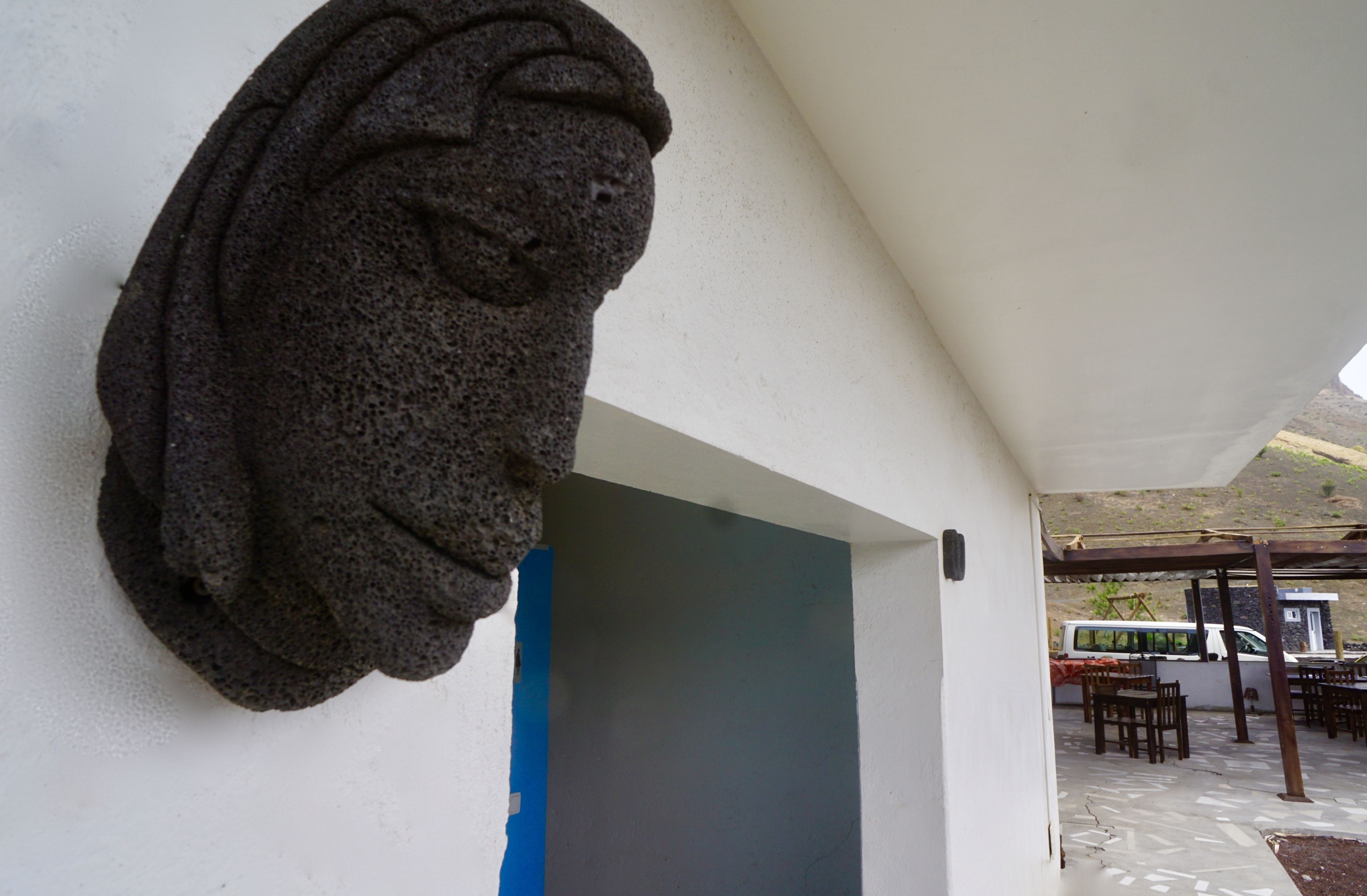 Escultura de cabeça de pedra em uma parede branca, porta azul e área de varanda externa com mesas e cadeiras.