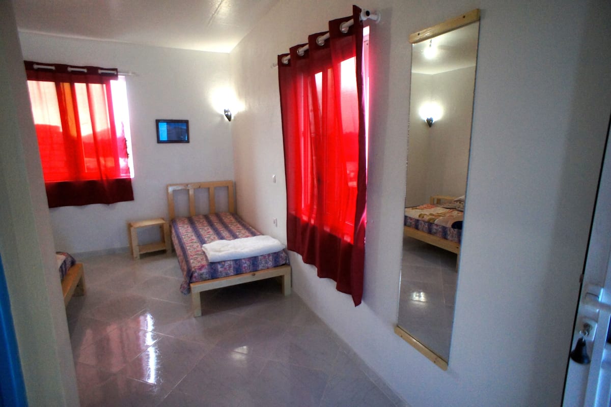 Quarto pequeno e bem arrumado, com uma cama de solteiro, mesa de cabeceira, espelho e cortinas vermelhas.