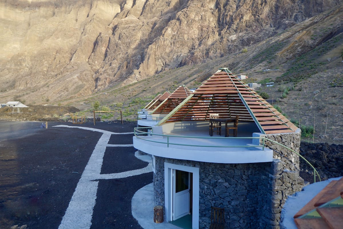 Um edifício moderno com base de pedra e telhado de madeira, localizado em área montanhosa com uma estrada bem visível à frente.