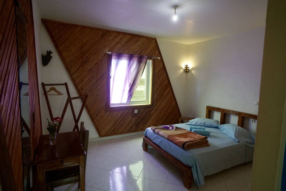 Um quarto com teto inclinado, uma cama com travesseiros e cobertor, uma mesa de madeira com um vaso de flores e uma janela com cortina roxa.
