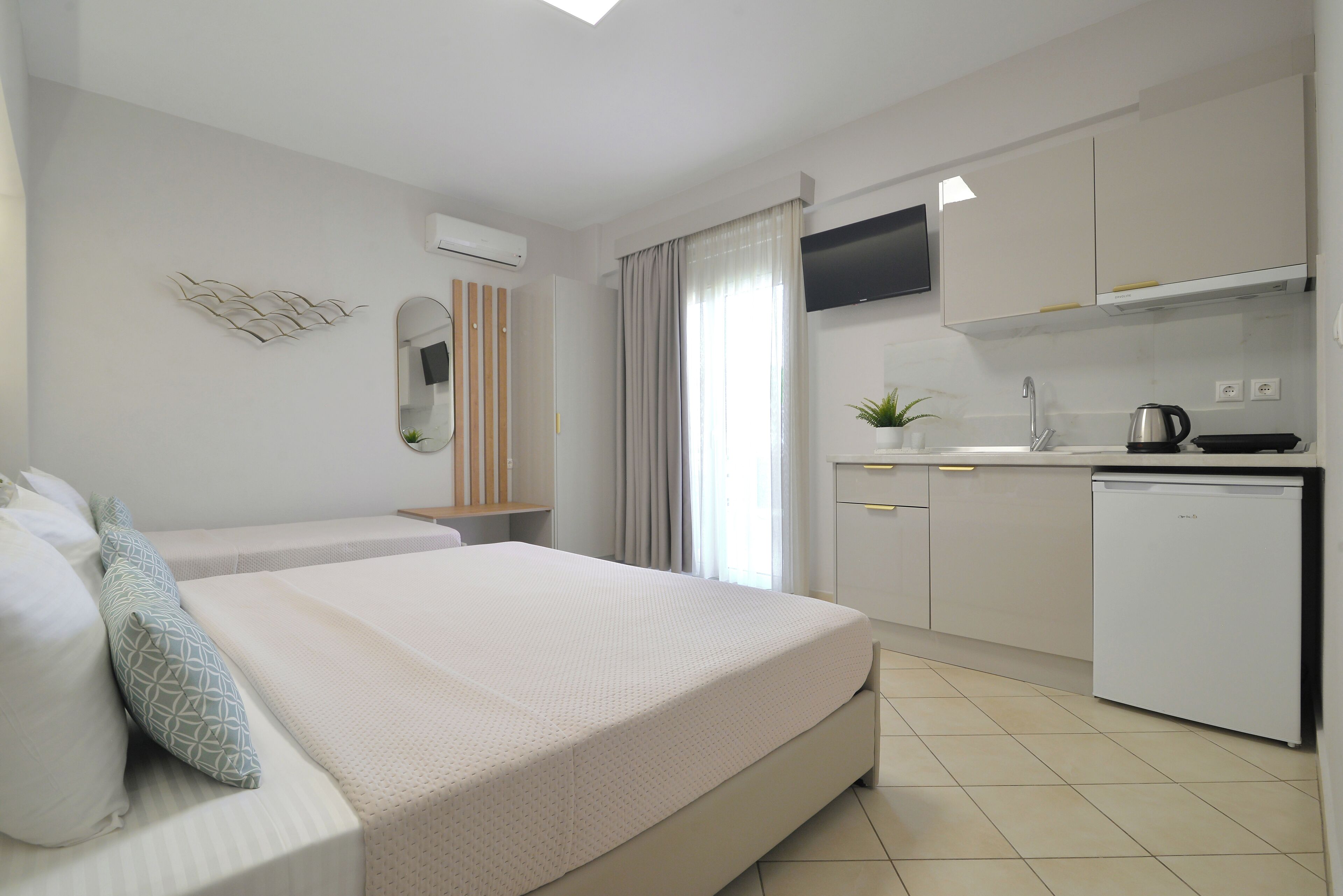 Quarto de hotel moderno com uma cama grande, uma pequena escrivaninha, uma cozinha compacta e uma televisão na parede.