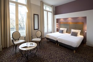 Quarto de hotel com duas camas, uma mesa redonda com um livro, duas cadeiras e uma janela grande com cortinas.
