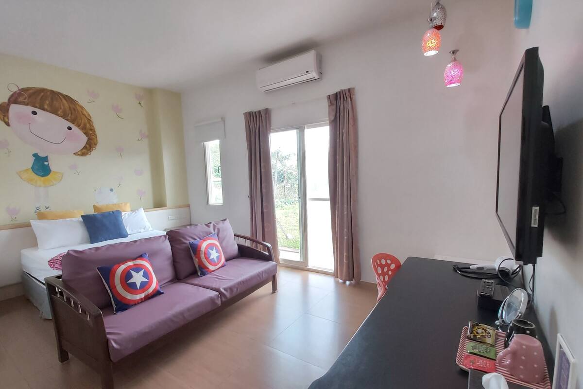 Um quarto aconchegante com um sofá decorado com almofadas do Capitão América, uma televisão de tela plana e uma janela grande com cortinas.