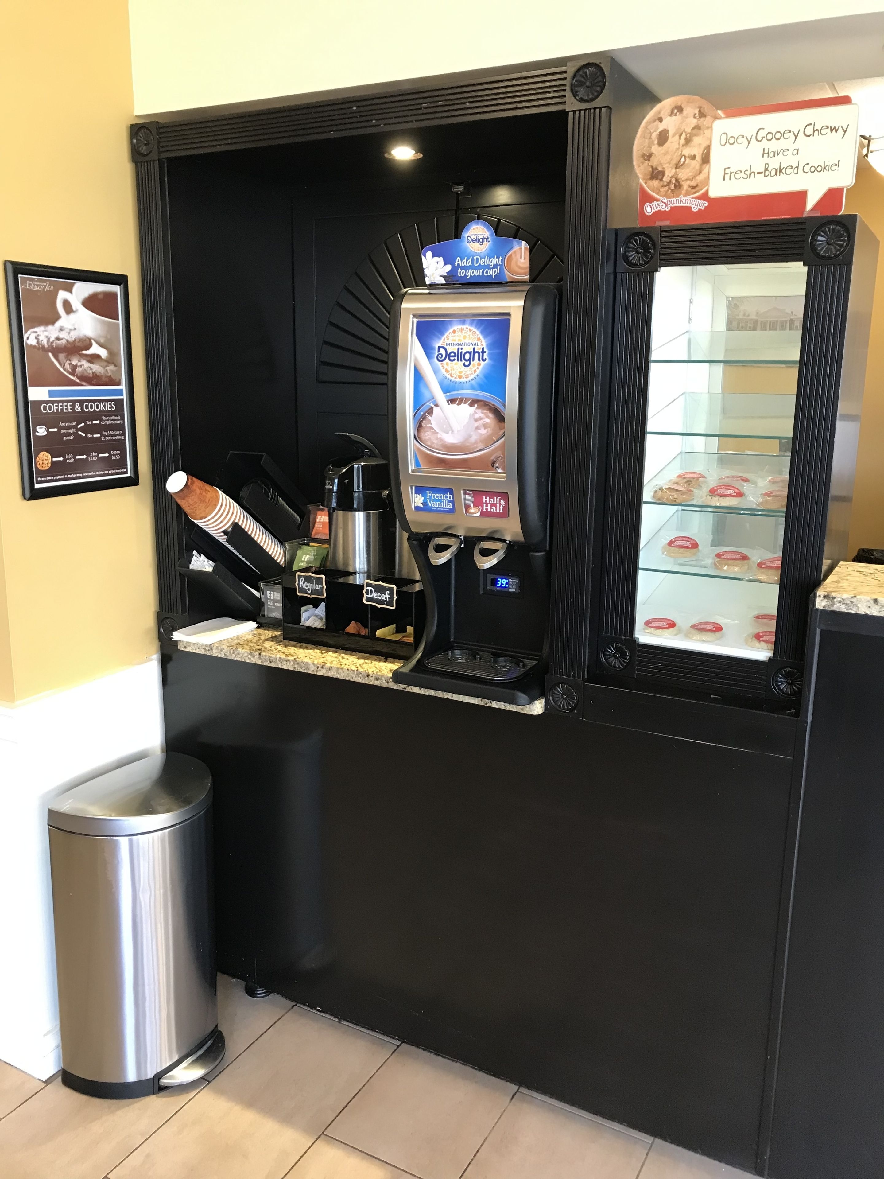 Máquina de café com vaporizador de leite, um cesto de lixo e uma vitrine com biscoitos.