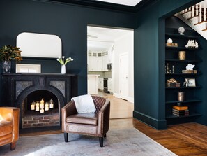 Um quarto com parede azul escura, lareira com velas acesas, poltrona de couro e estante com objetos decorativos.