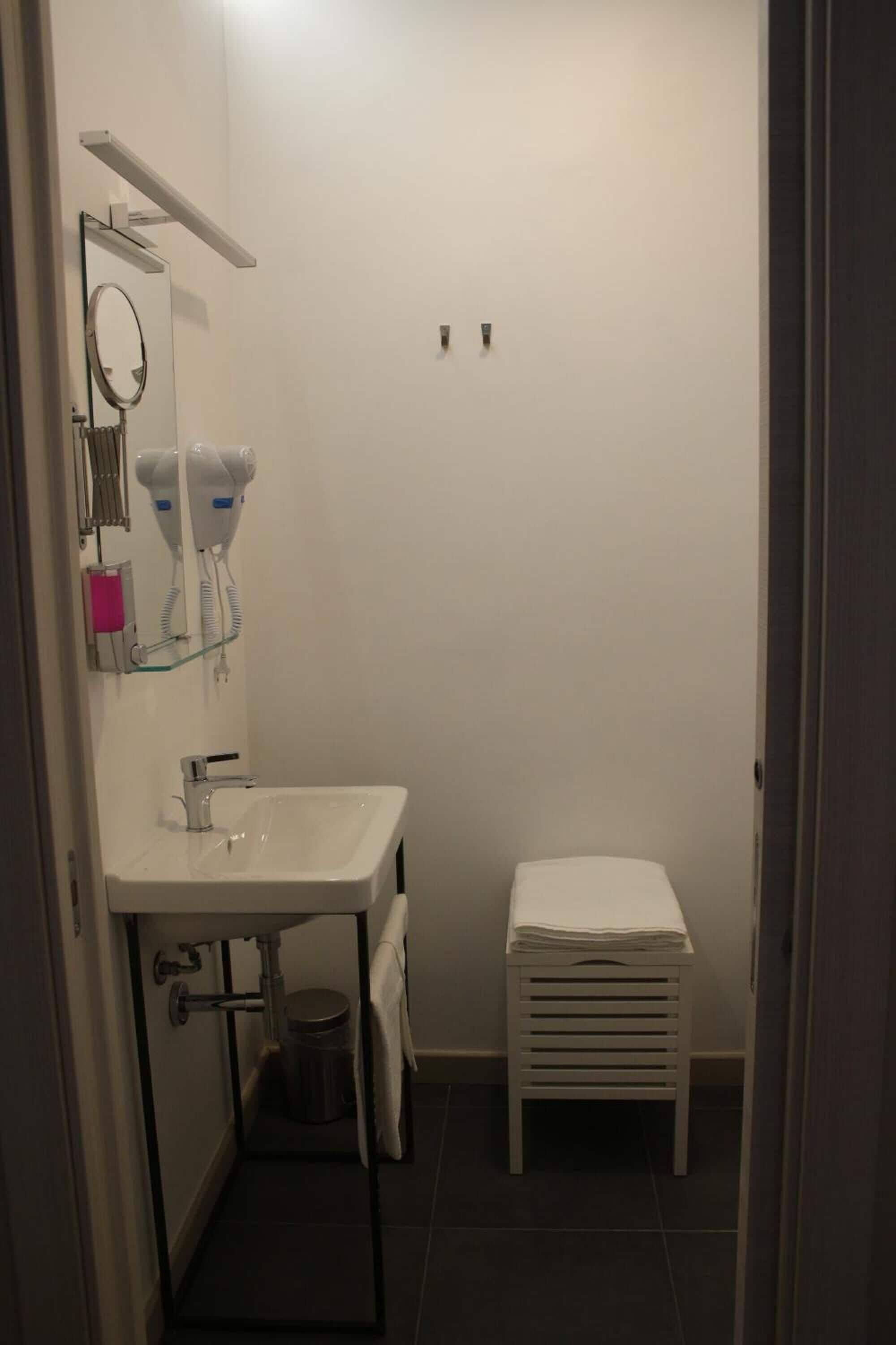 Casa de banho pequena e compacta com lavatório branco, espelho e secador de cabelo. Há um banquinho pequeno e um cesto de lixo.