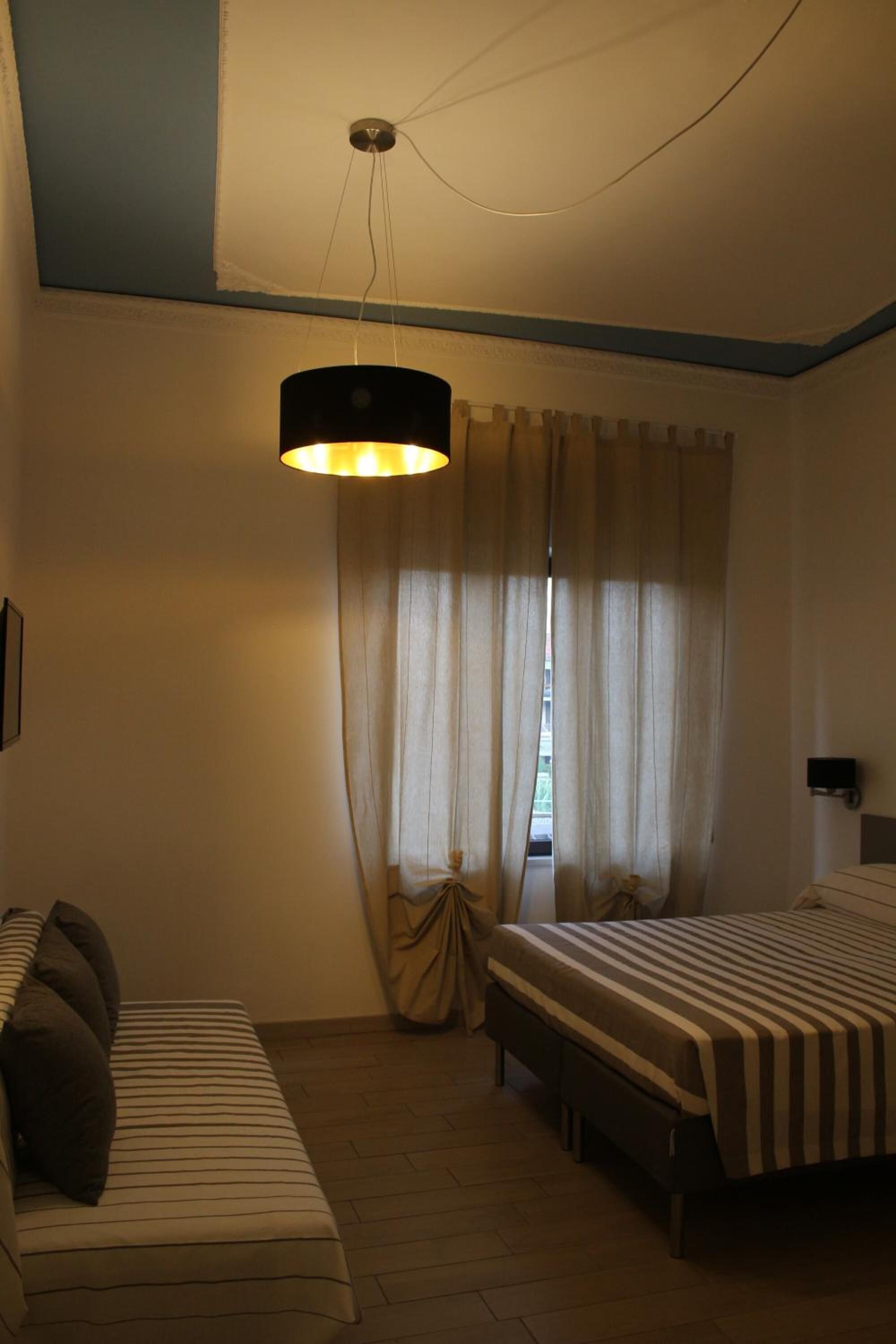 Quarto de hotel com cama, sofá, luminária pendente e janela com cortinas.