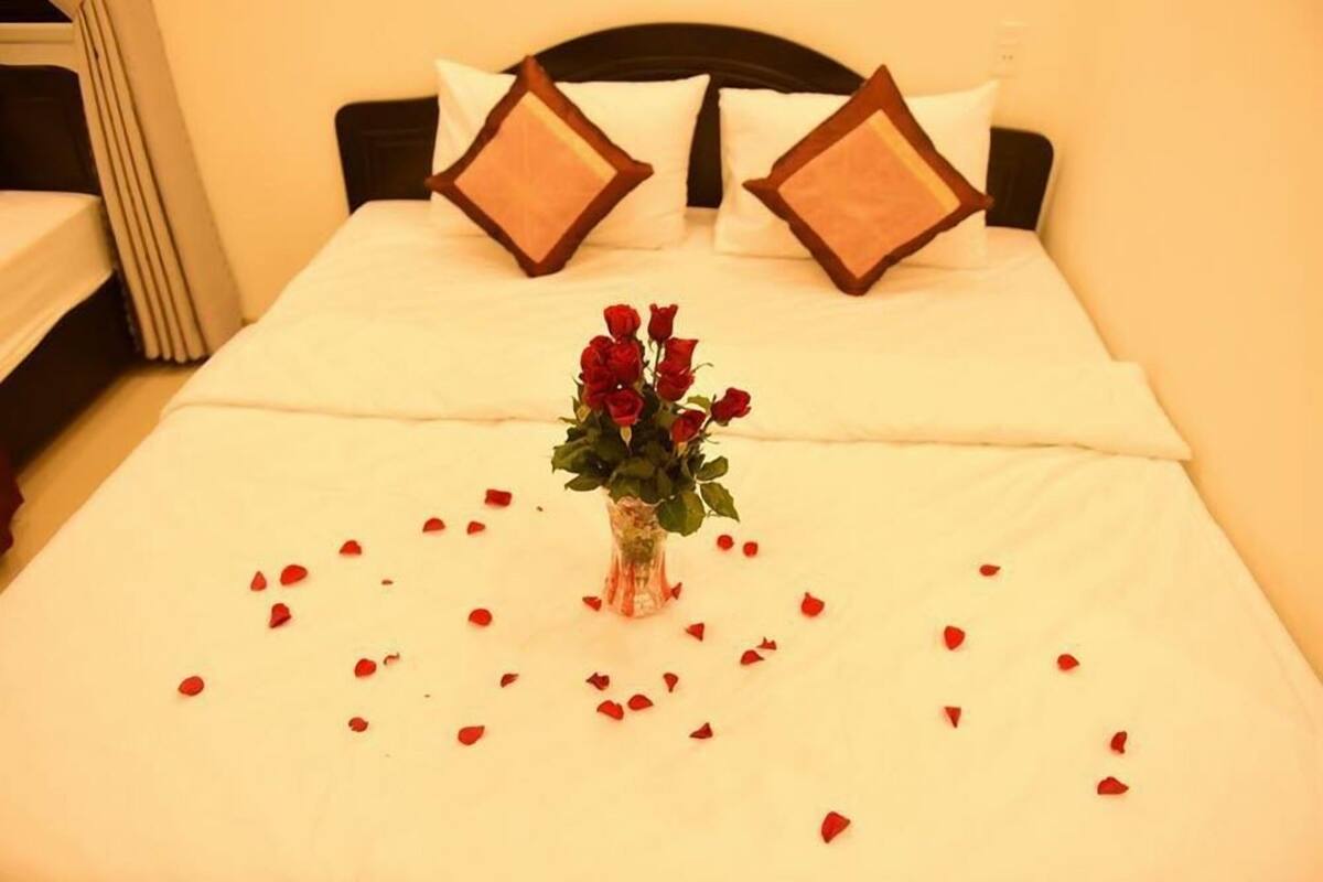 Uma cama com roupa de cama branca, duas almofadas castanhas e um vaso com rosas vermelhas no centro.