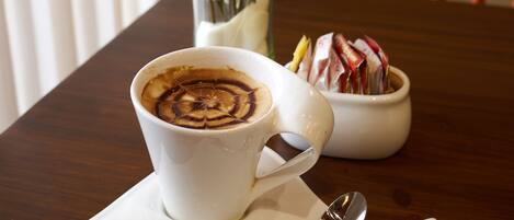 Uma chávena de café com arte de latte numa bandeja branca, uma colher, um pequeno vaso com flores cor-de-rosa e uma caixa de lenços sobre uma mesa de madeira.