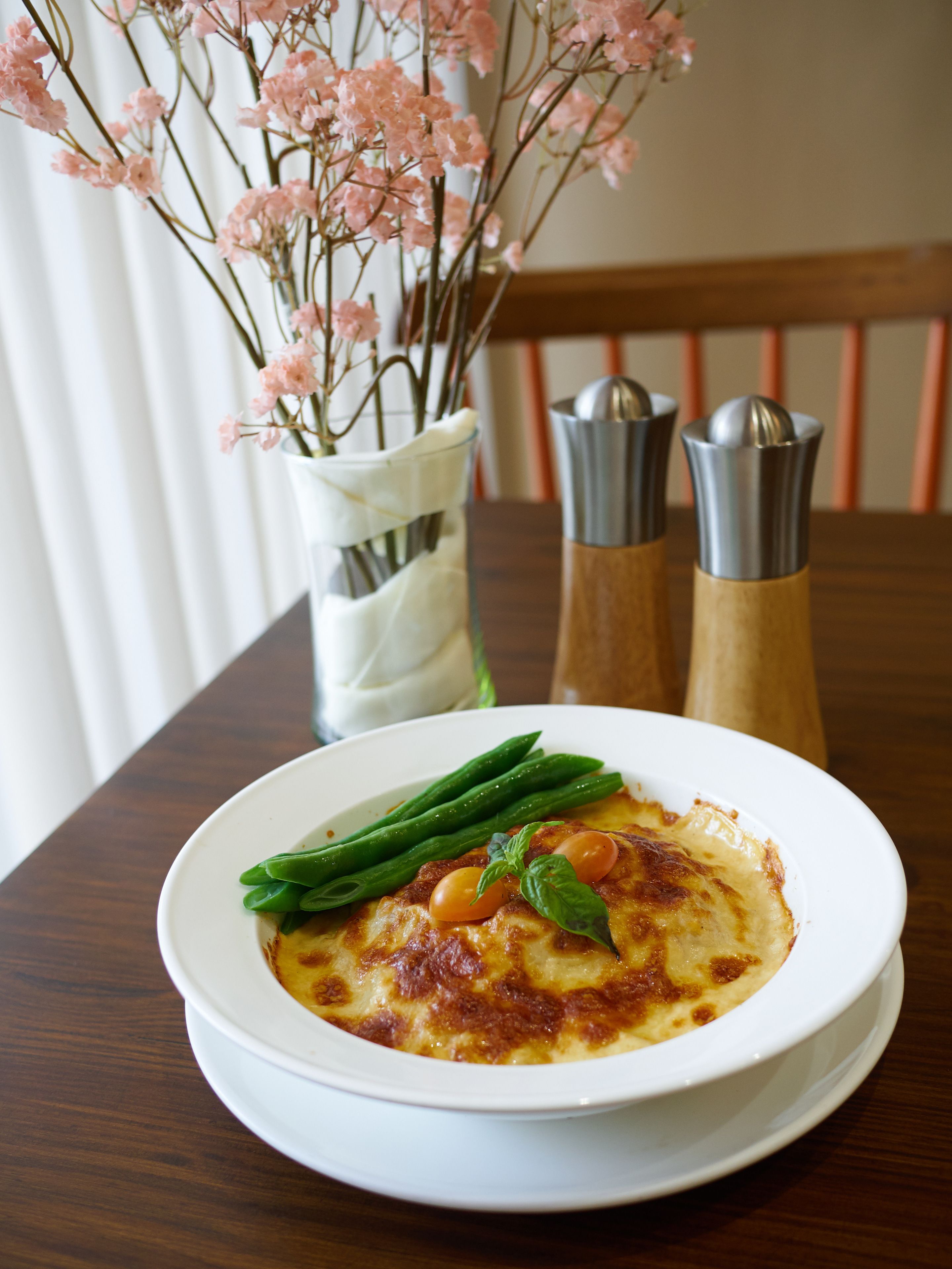 Uma tigela branca com comida sobre uma mesa de madeira, com um vaso de flores cor-de-rosa e dois saleiros e pimenteiros ao fundo.