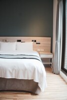 Quarto moderno com cabeceira de madeira, cama com roupa de cama branca, mesa de cabeceira e janela com cortinas.
