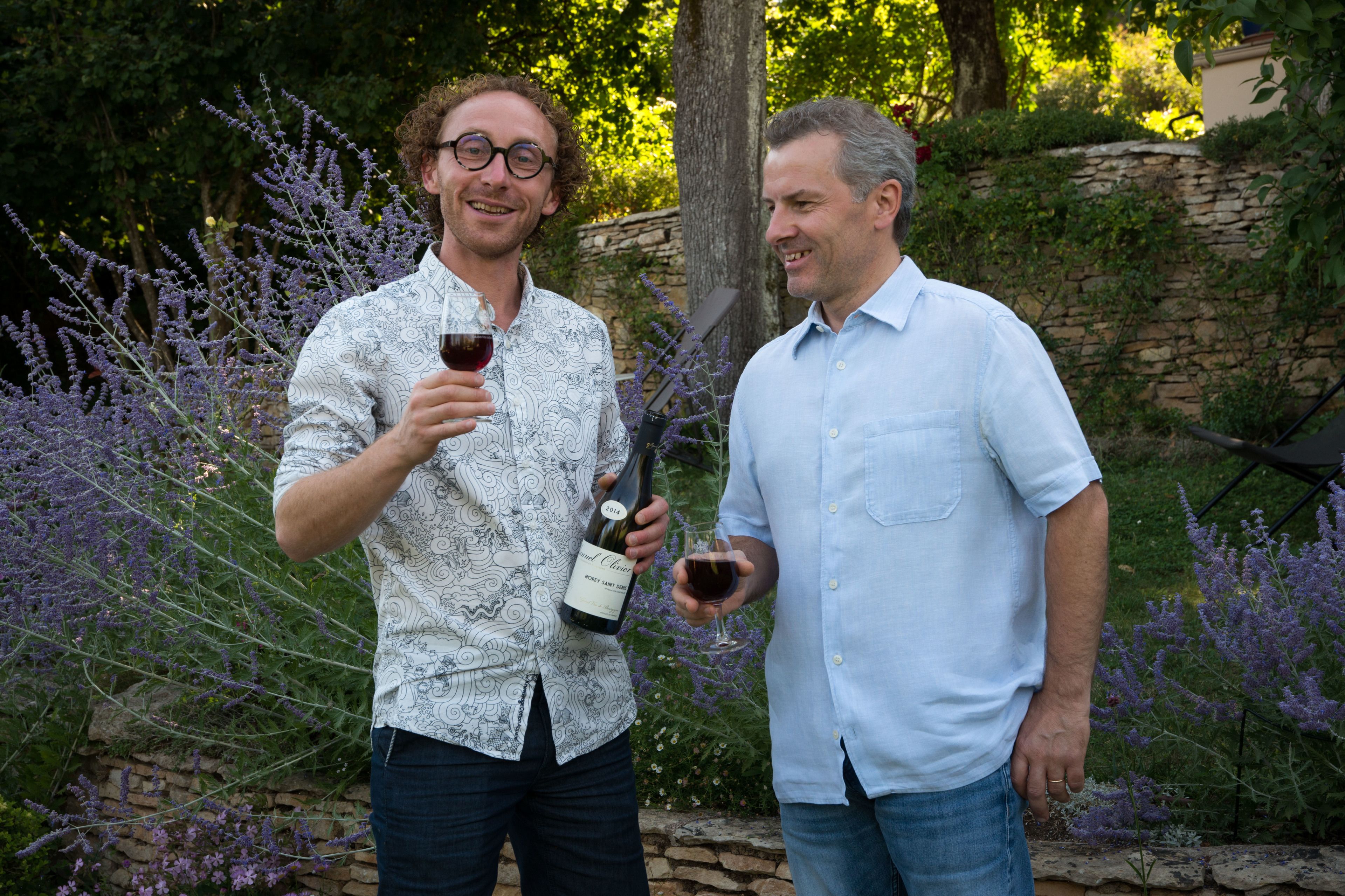 Duas pessoas segurando garrafas de vinho e copos num jardim com plantas de lavanda.