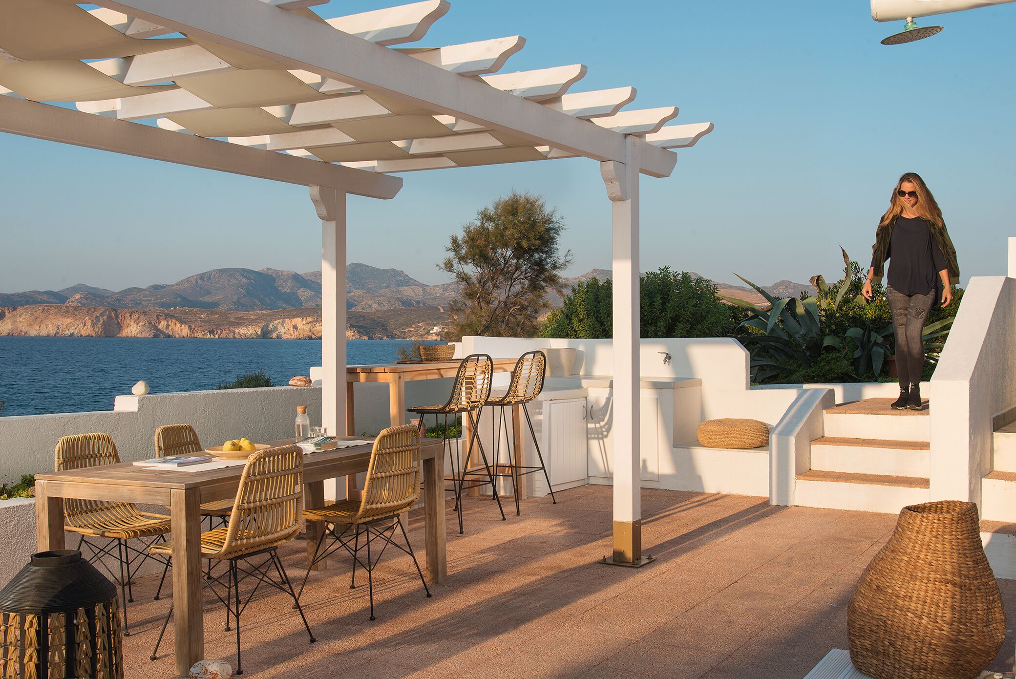 Um pátio externo com mesa e cadeiras, uma pérgola e vista para o mar e montanhas.
