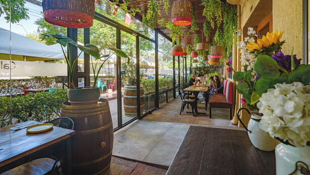 Uma área de refeições ao ar livre com mesas de madeira, plantas penduradas e decoração rústica.