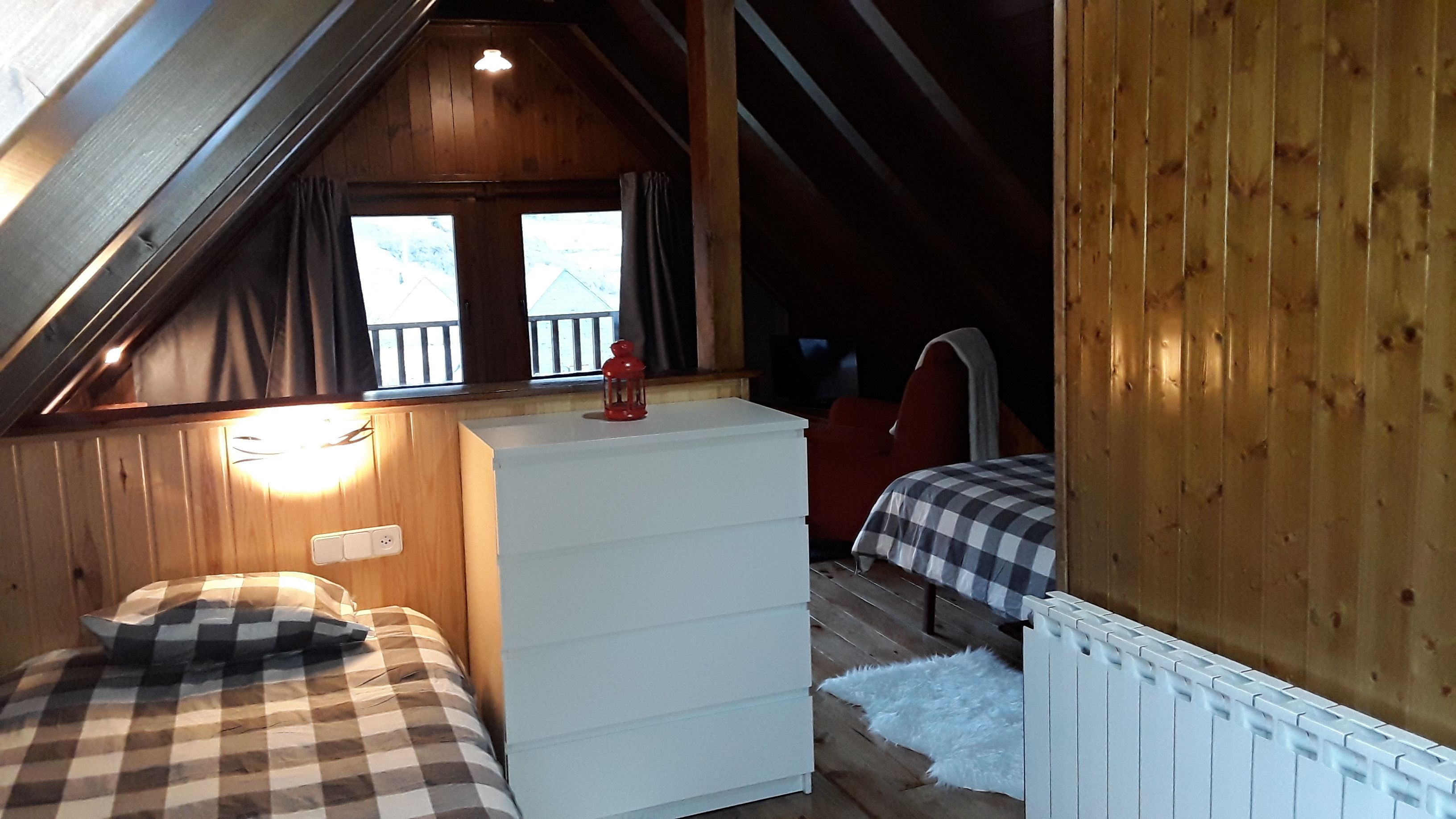 Um quarto de sótão acolhedor, com teto inclinado, uma cômoda branca, duas camas com colchas xadrez e vista para o exterior através de uma janela.