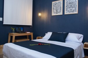 Uma cama bem arrumada com colcha azul escura e almofadas brancas, uma mesa de cabeceira com abajur, e duas obras de arte emolduradas na parede.