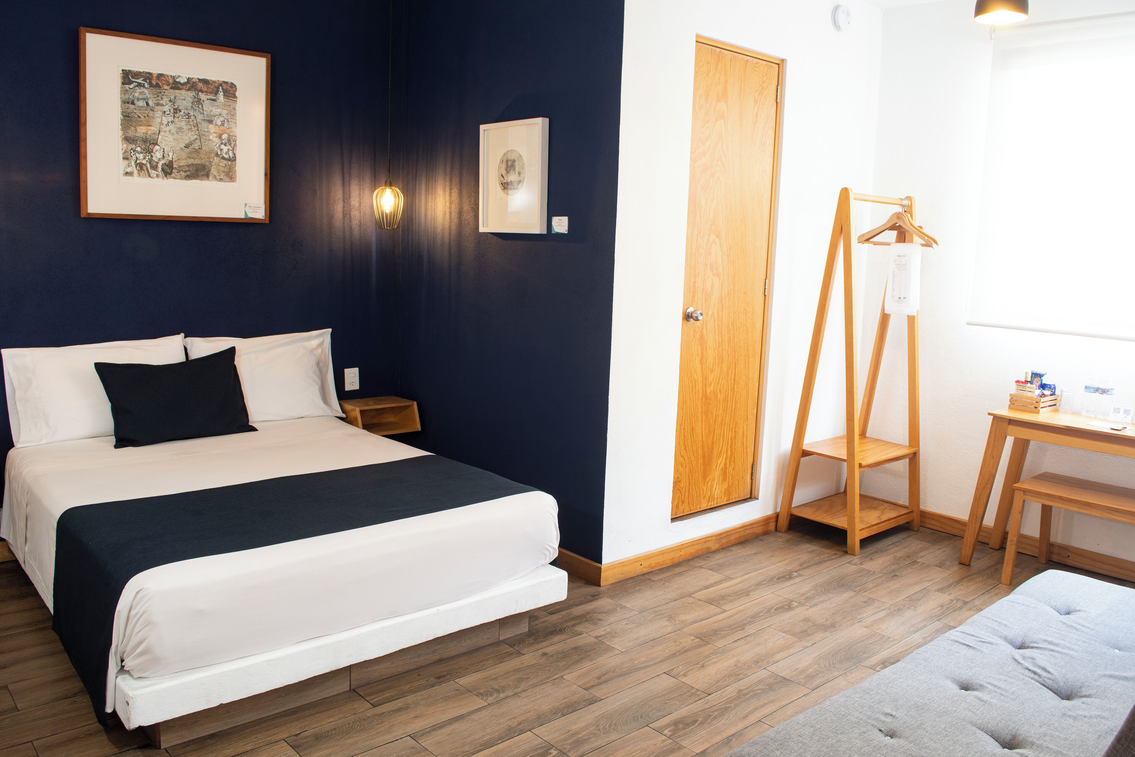 Quarto com cama, penteadeira de madeira, escrivaninha com cabideiro e um quadro na parede.